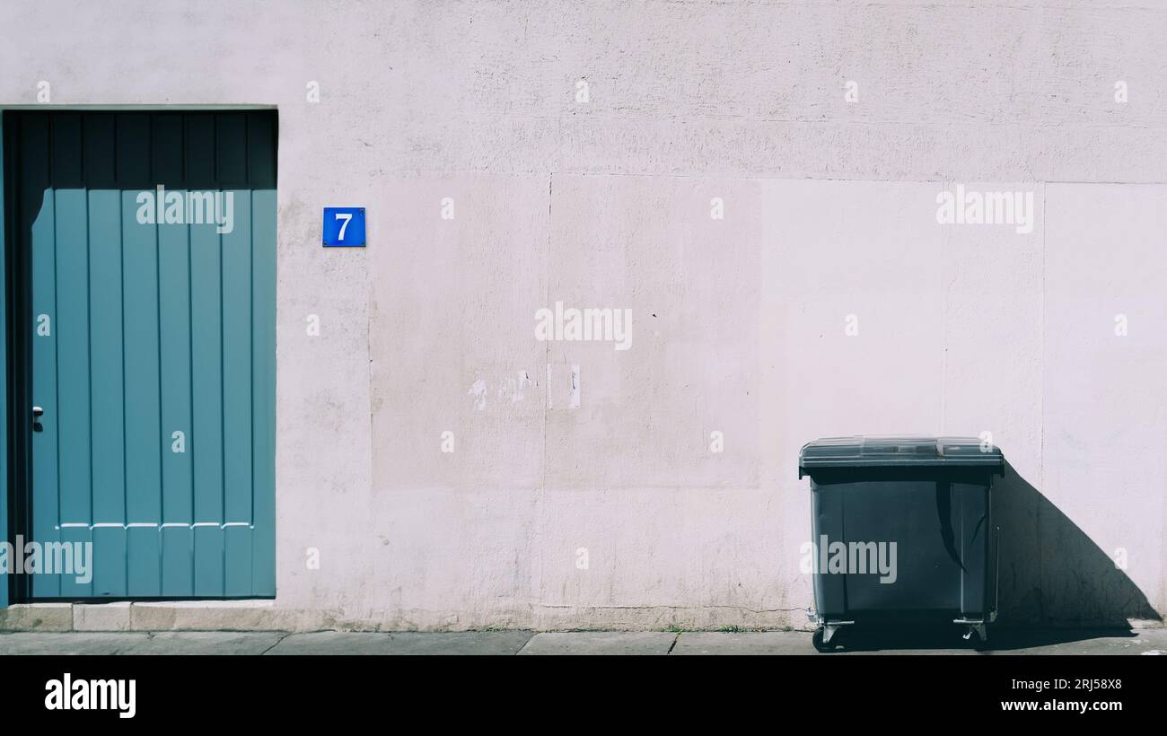 A trash bin in front of a building wall with a blue door illuminated by ...