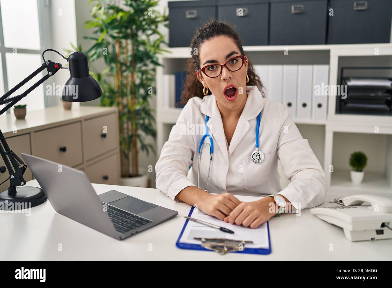 Young hispanic woman wearing doctor uniform and stethoscope in shock ...