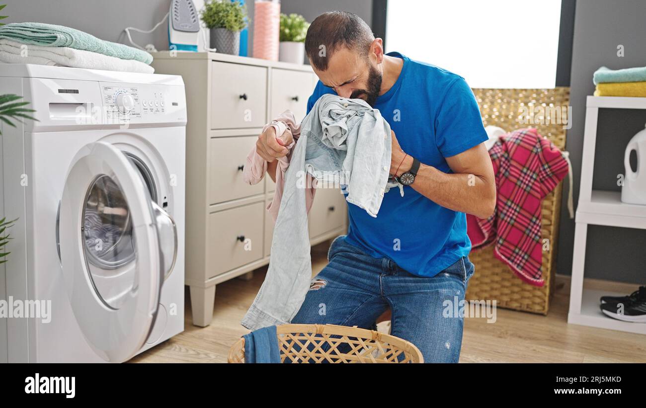 Young hispanic man washing clothes smelling dirty clothes at laundry ...