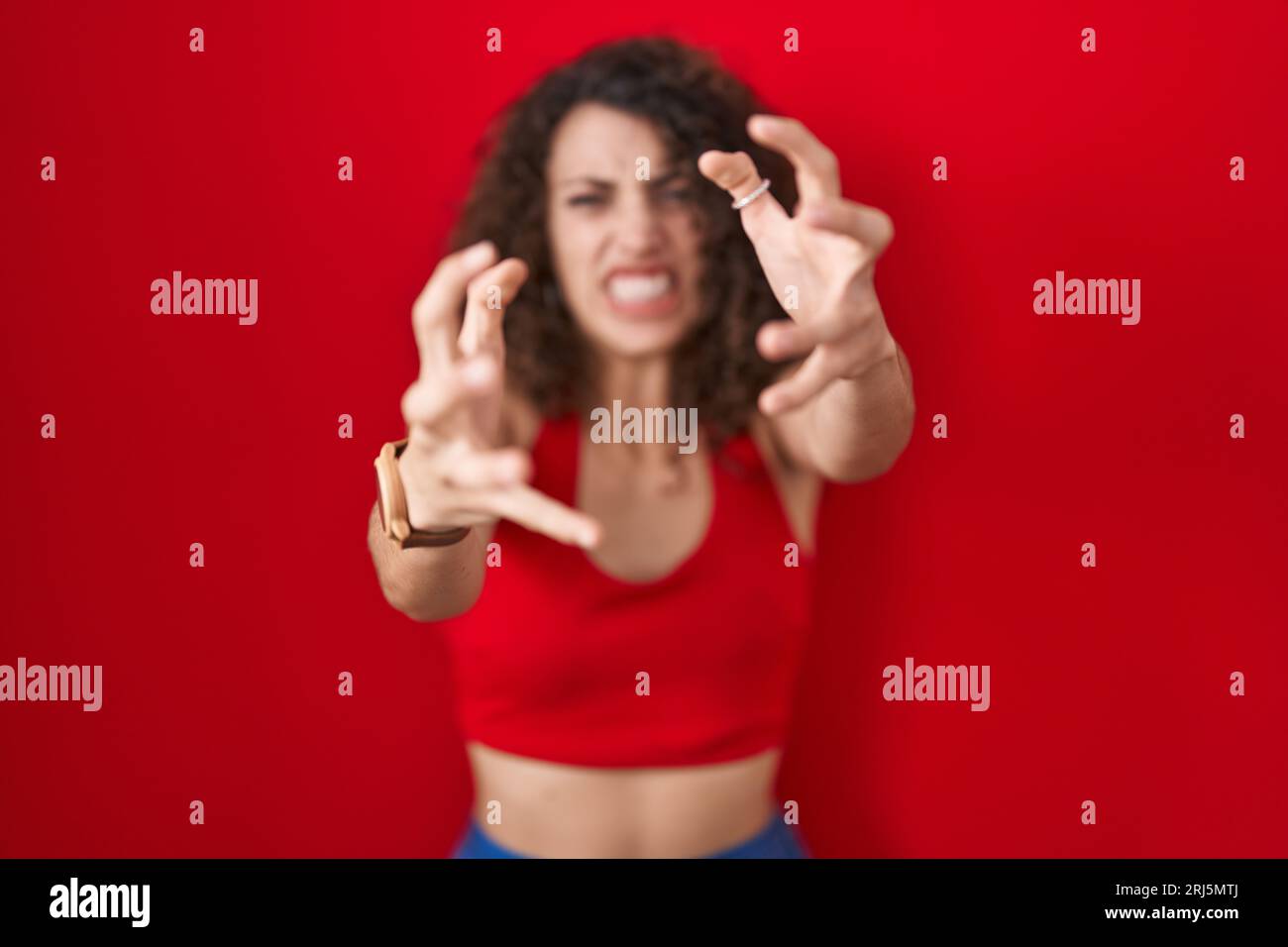 Hispanic woman with curly hair standing over red background shouting ...