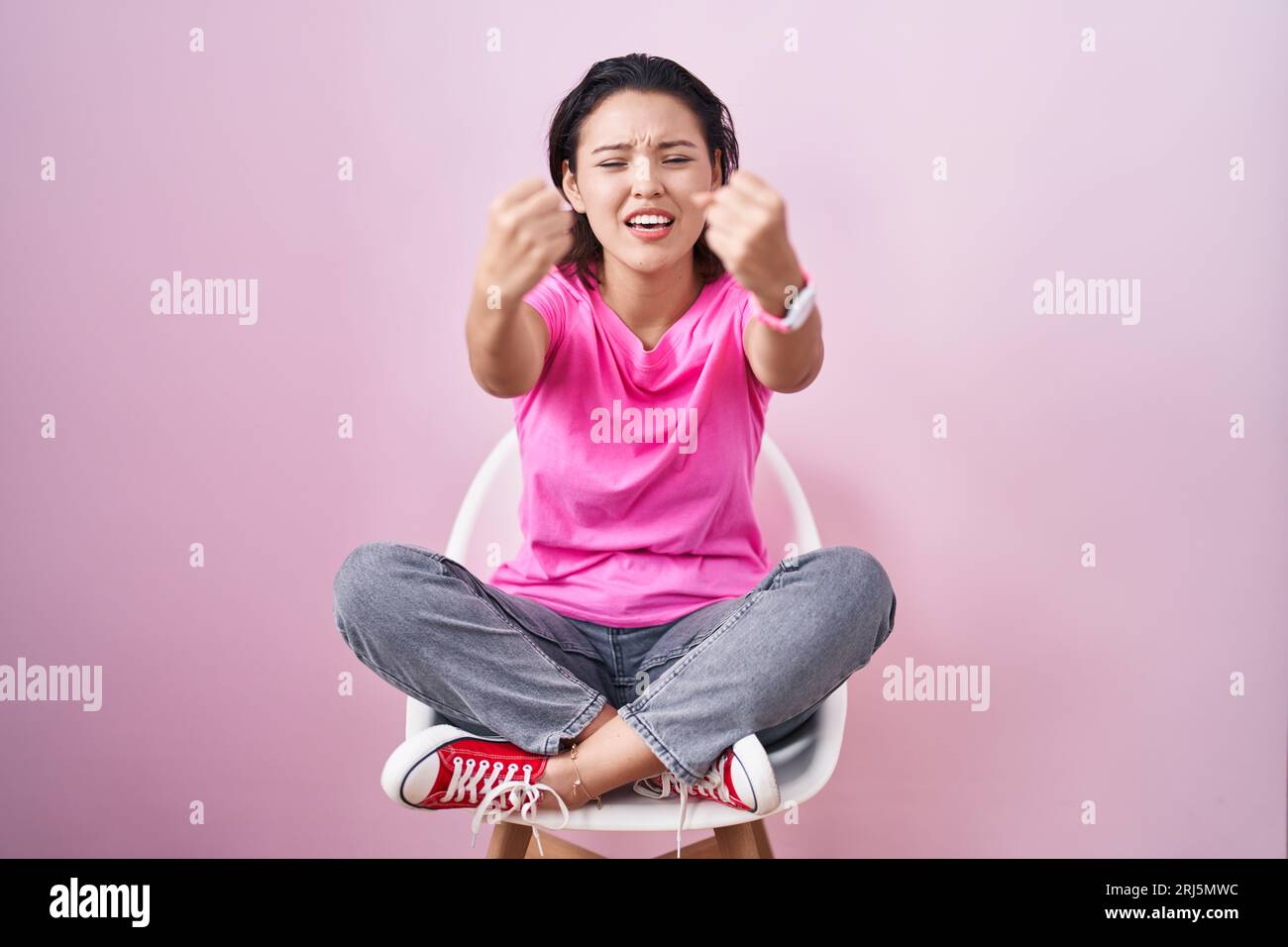 Hispanic young woman sitting on chair over pink background angry and ...