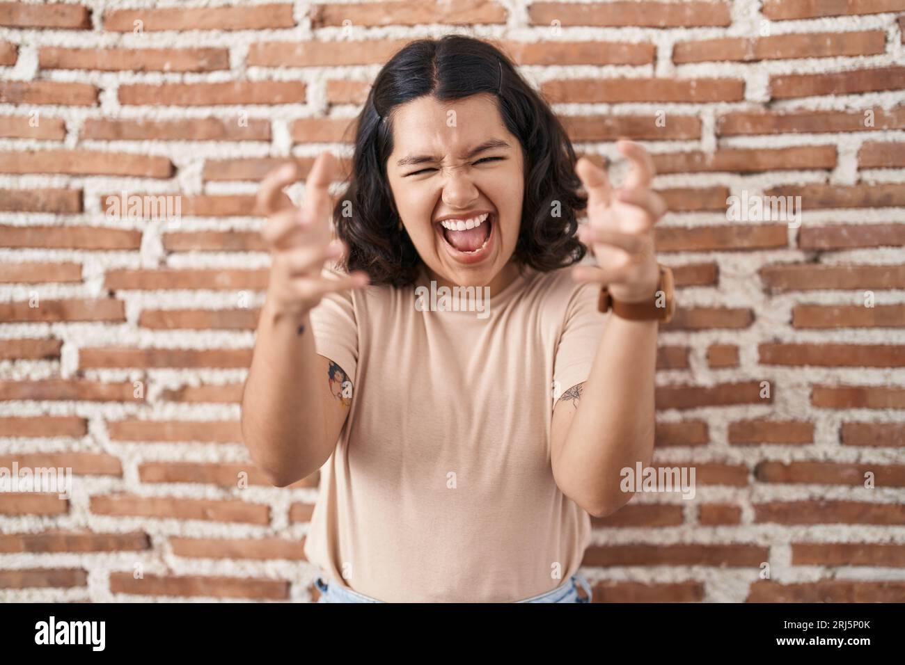 Young hispanic woman standing over bricks wall shouting frustrated with ...