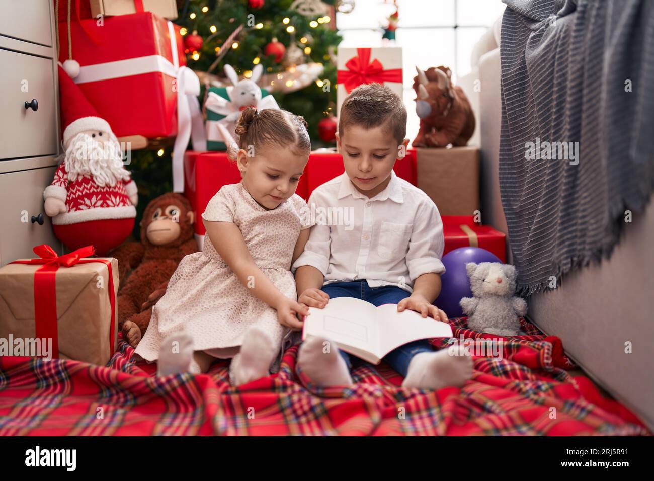 Two kids reading book sitting on floor by christmas tree at home Stock ...