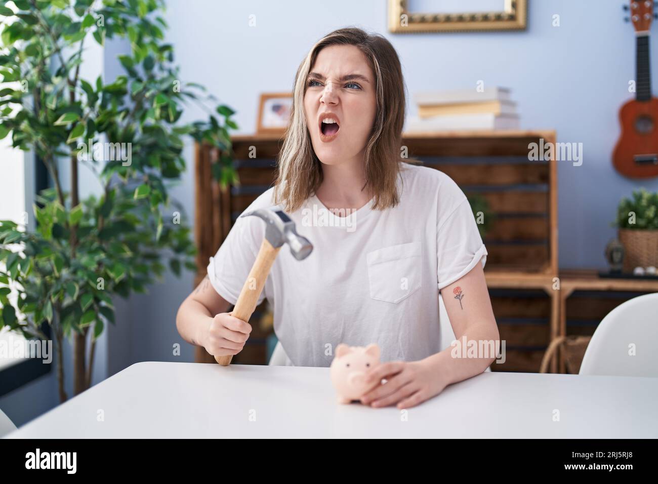 Blonde caucasian woman holding piggy bank and hammer angry and mad ...
