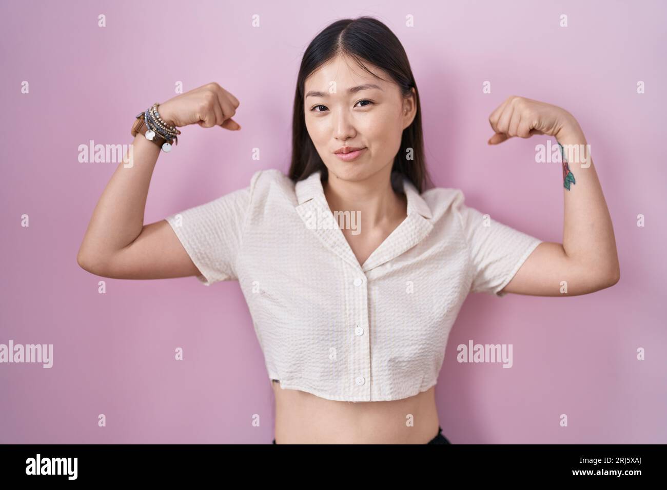 Chinese young woman standing over pink background showing arms muscles ...