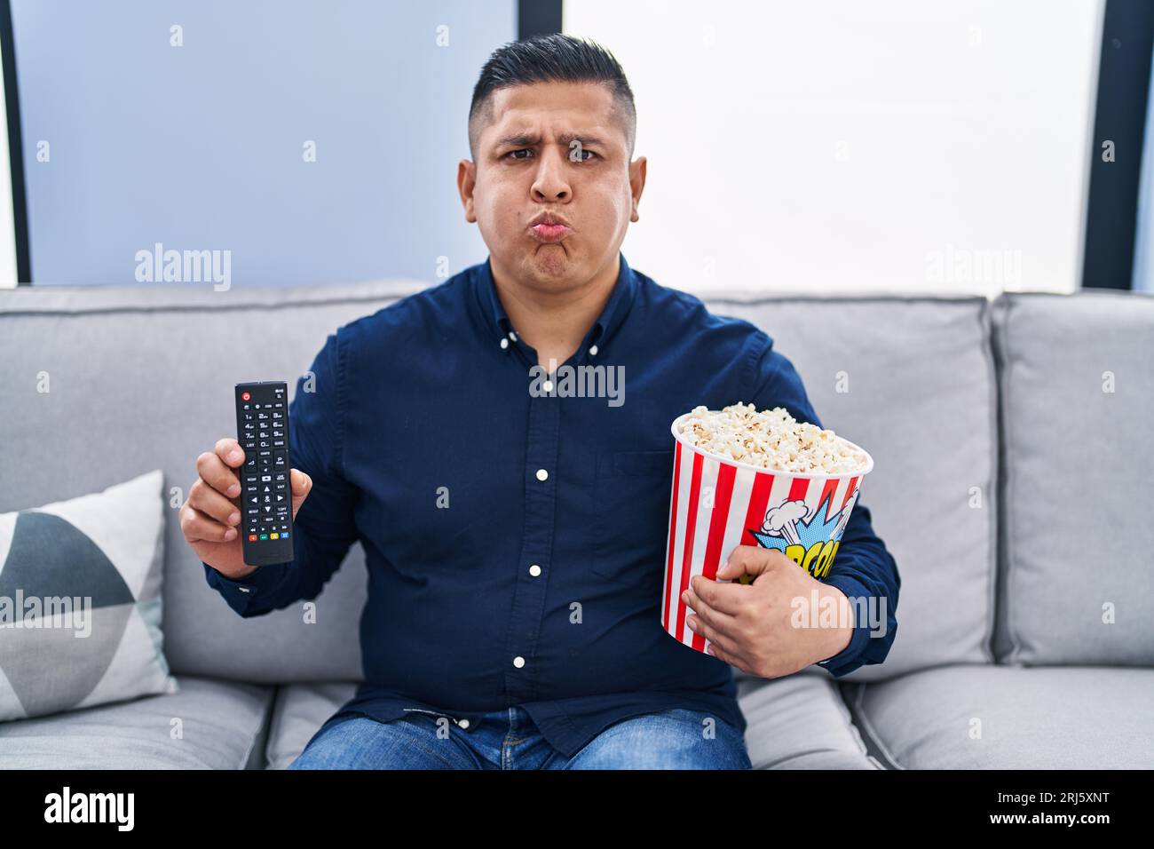 Hispanic young man eating popcorn using tv control puffing cheeks with ...