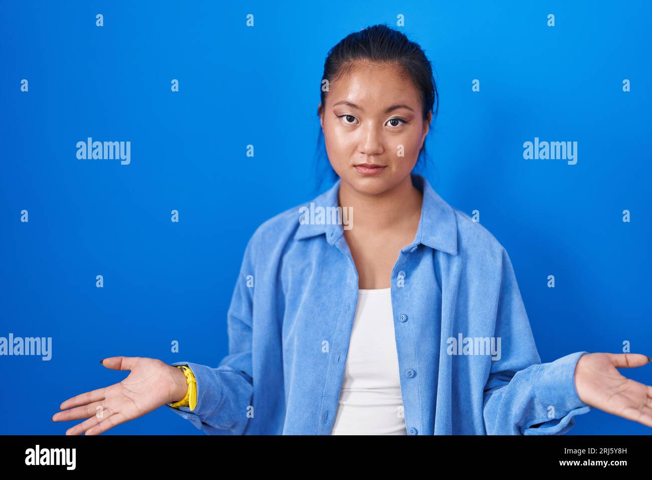 Asian young woman standing over blue background clueless and confused ...