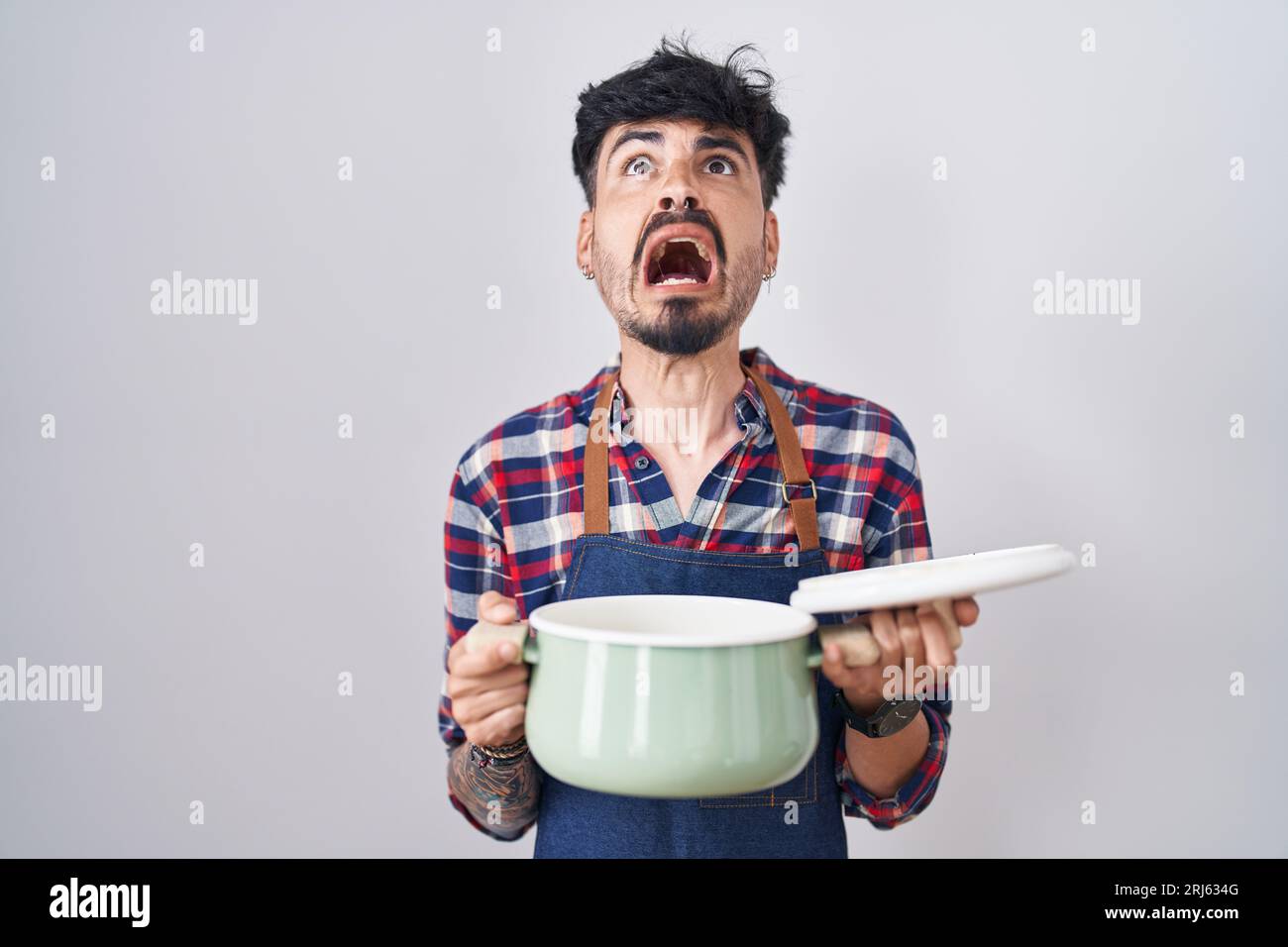 Young hispanic man with beard wearing apron holding cooking pot angry ...