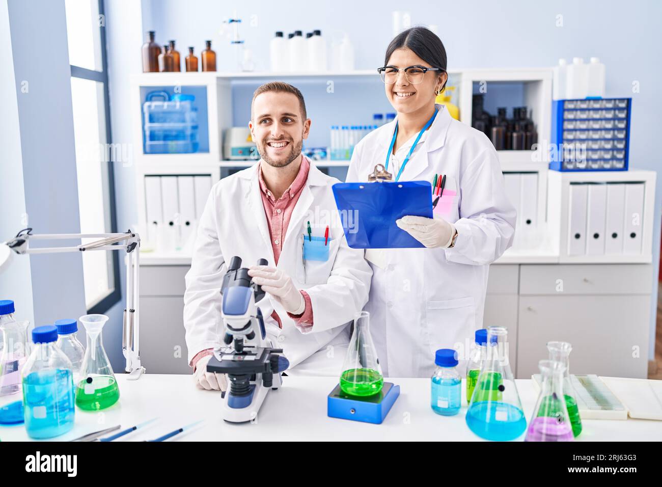 Two young people working at scientist laboratory looking positive and ...