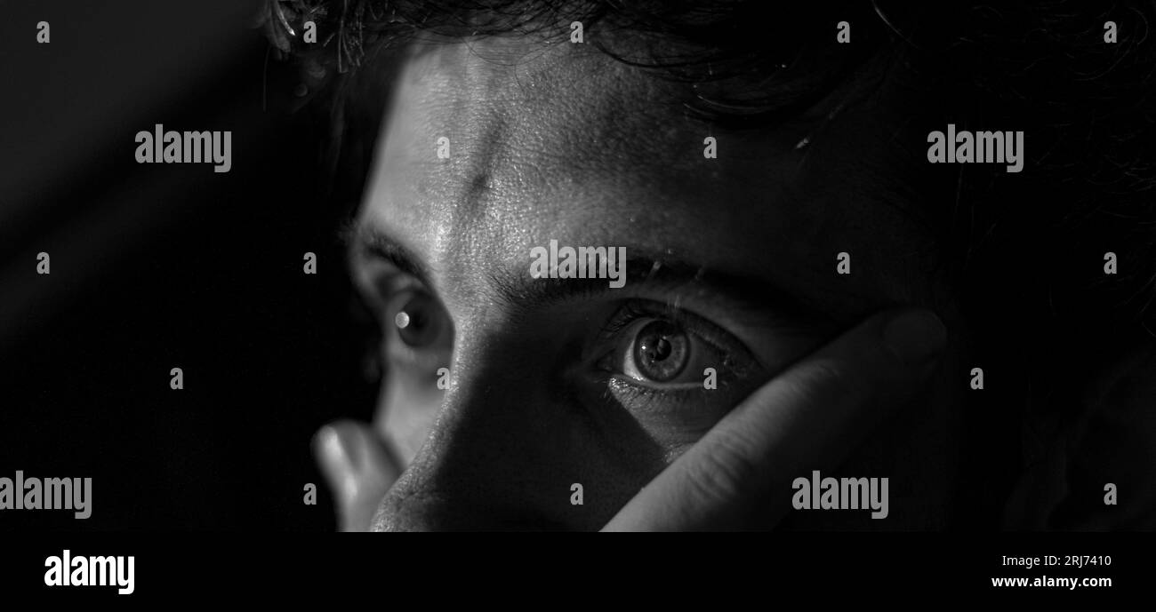 A grayscale closeup of the sad eyes of a young man in a dark ...