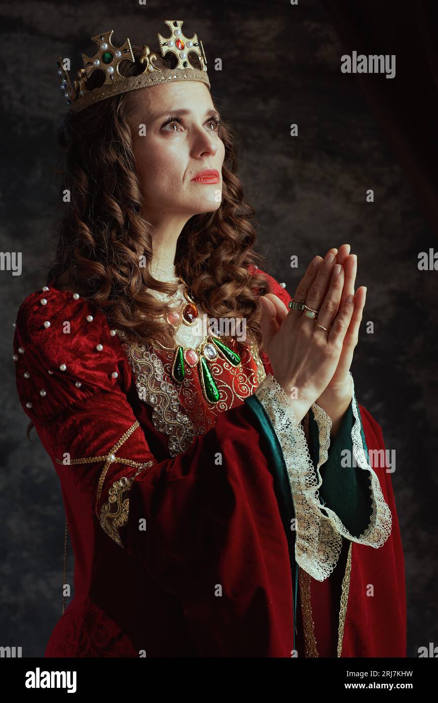 medieval queen in red dress with crown praying on dark gray background ...