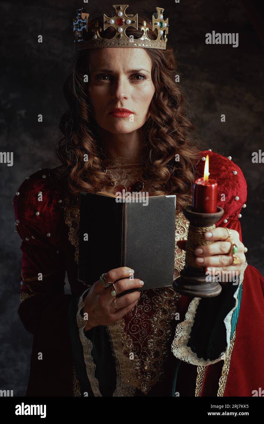 medieval queen in red dress with book, candle and crown on dark gray ...