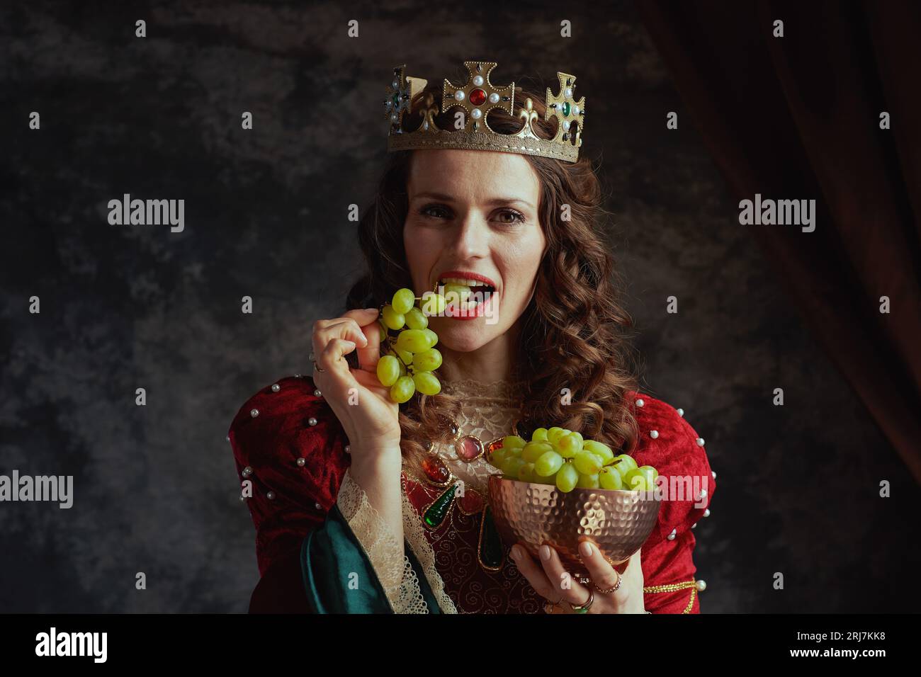 medieval queen in red dress with plate of grapes and crown on dark gray ...