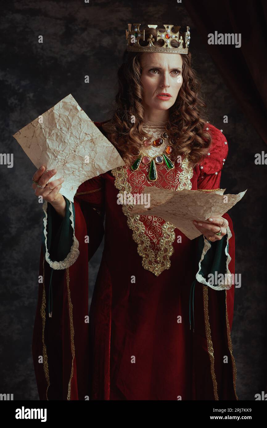 unhappy medieval queen in red dress with parchment and crown on dark ...
