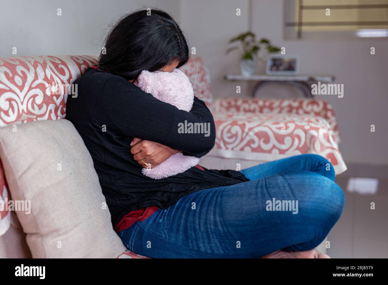 A sad woman sits on a couch, tightly hugging a pillow. She looks lost ...