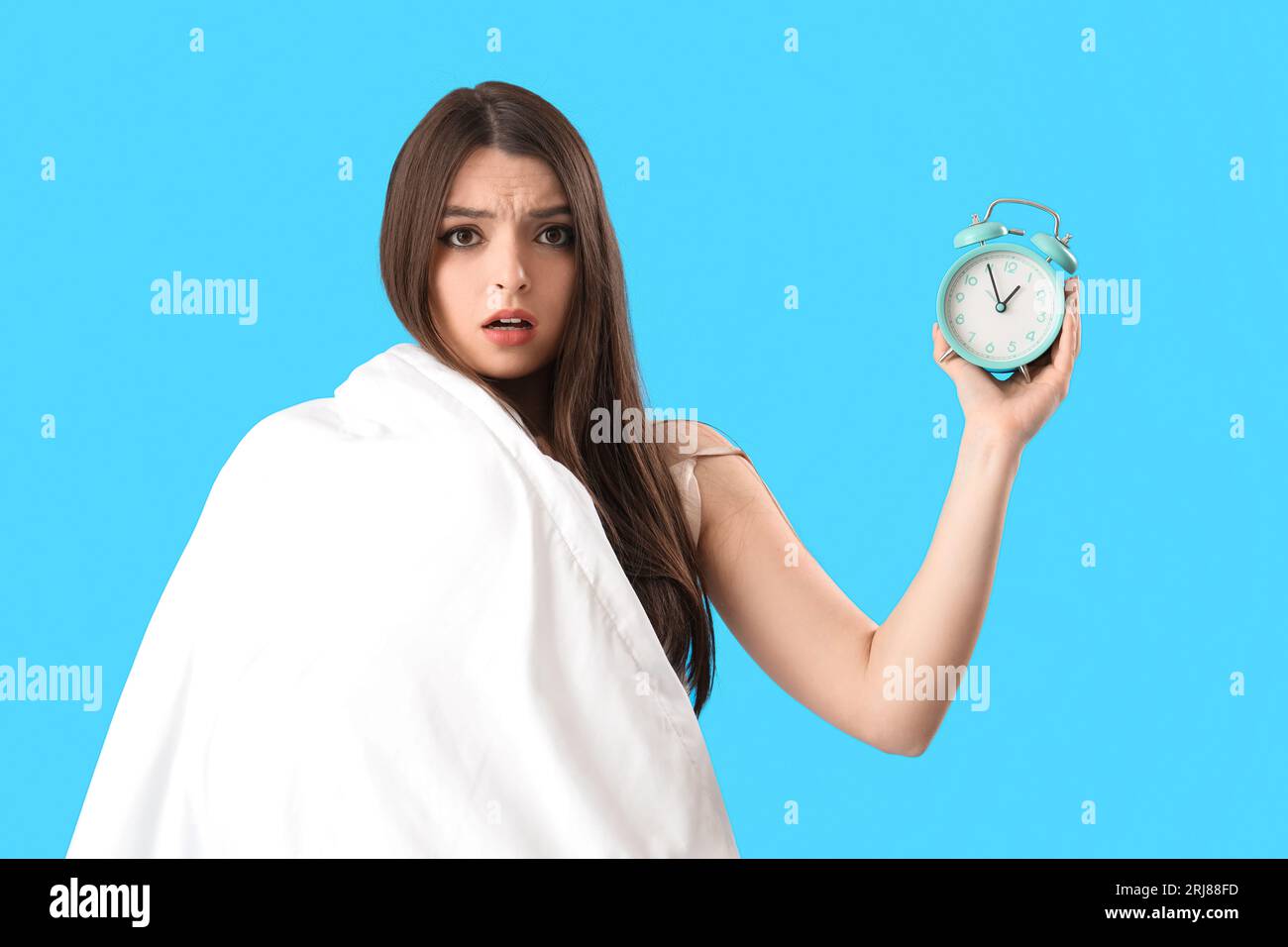 Upset young woman with alarm clock and soft blanket on blue background ...