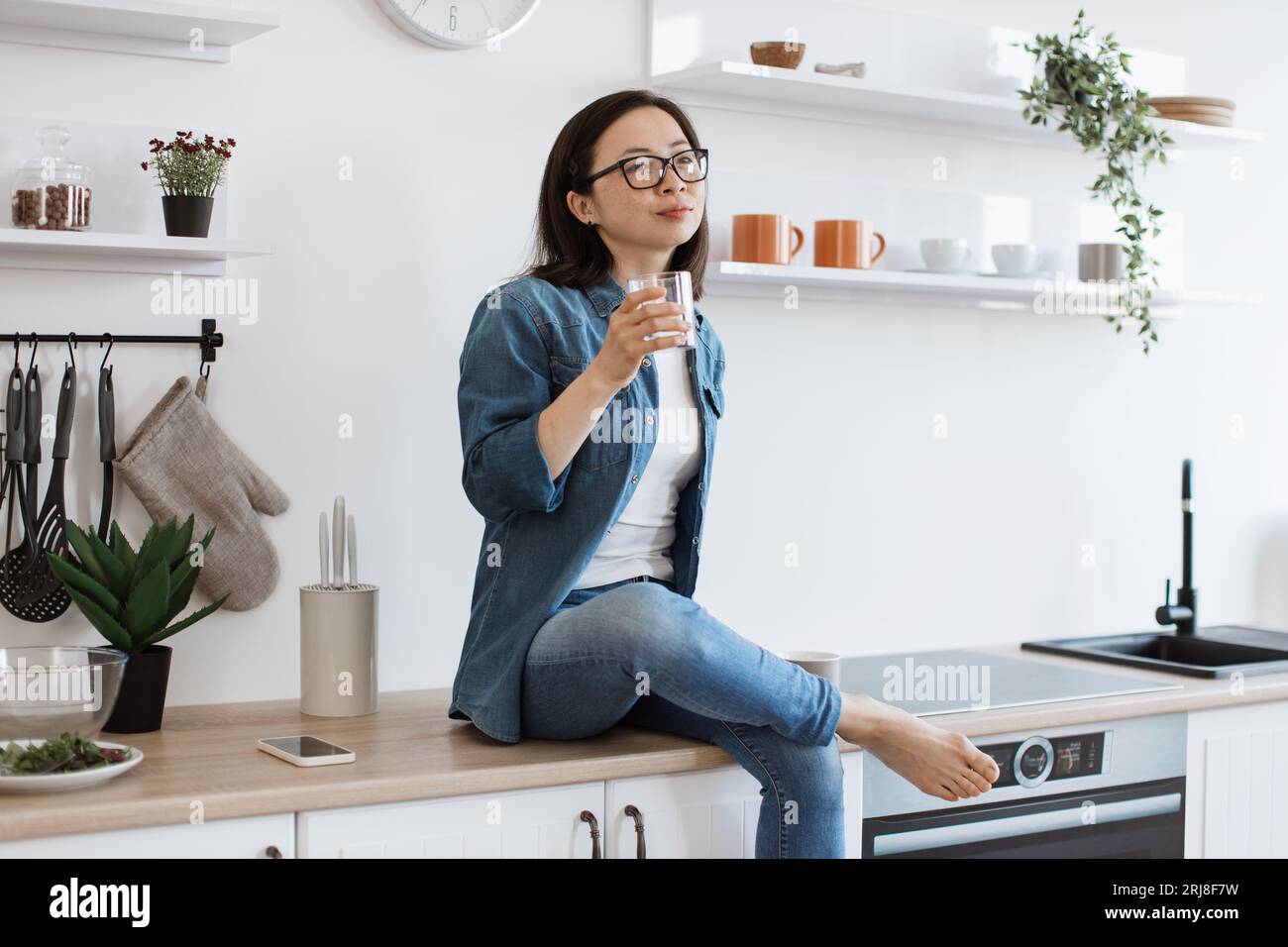 Relaxed barefoot lady in jeans daydreaming with glass full of liquid ...