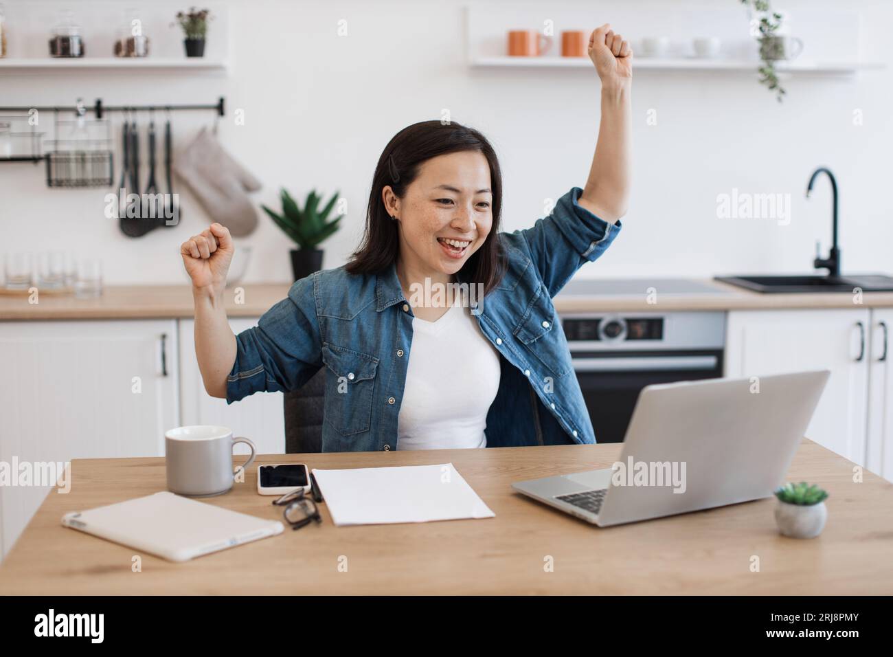Thrilled asian woman raising hands in winner's gesture while looking at digital screen of ...
