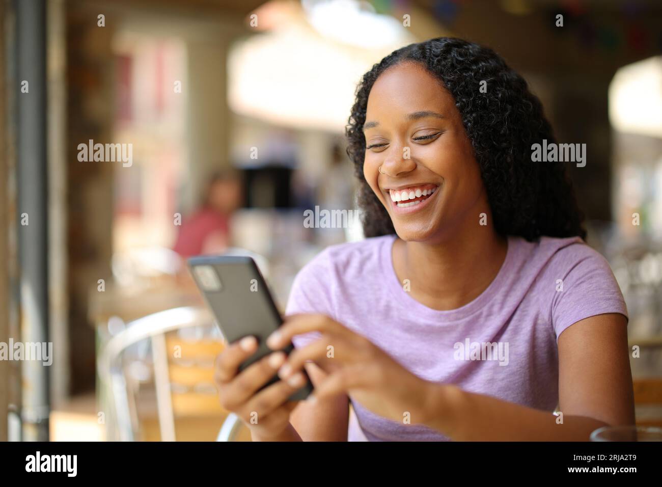 Happy black woman using cell phone laughing in a coffee shop terrace ...