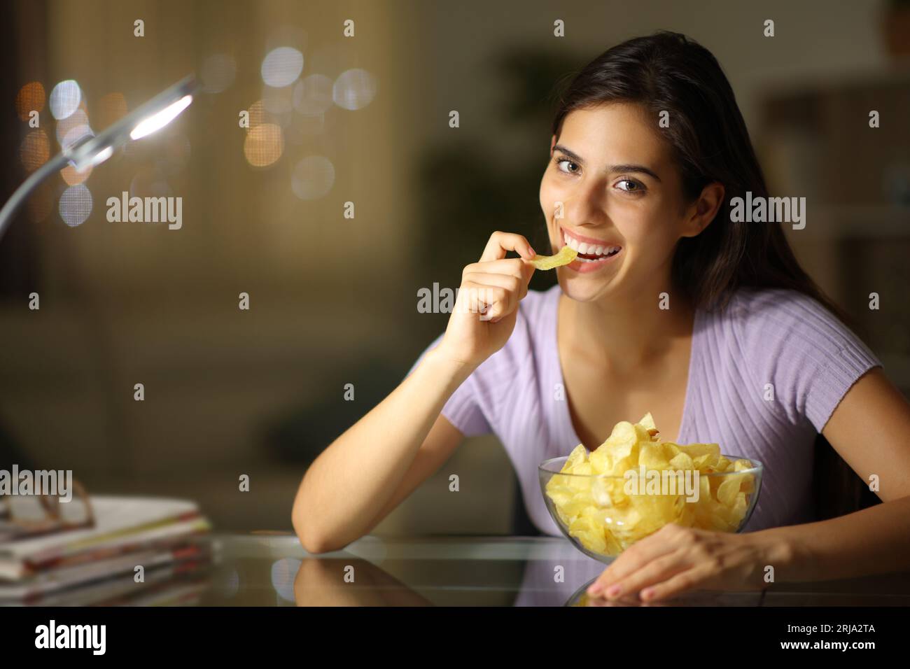 Happy woman in the night eating potato chips looking at camera at home ...