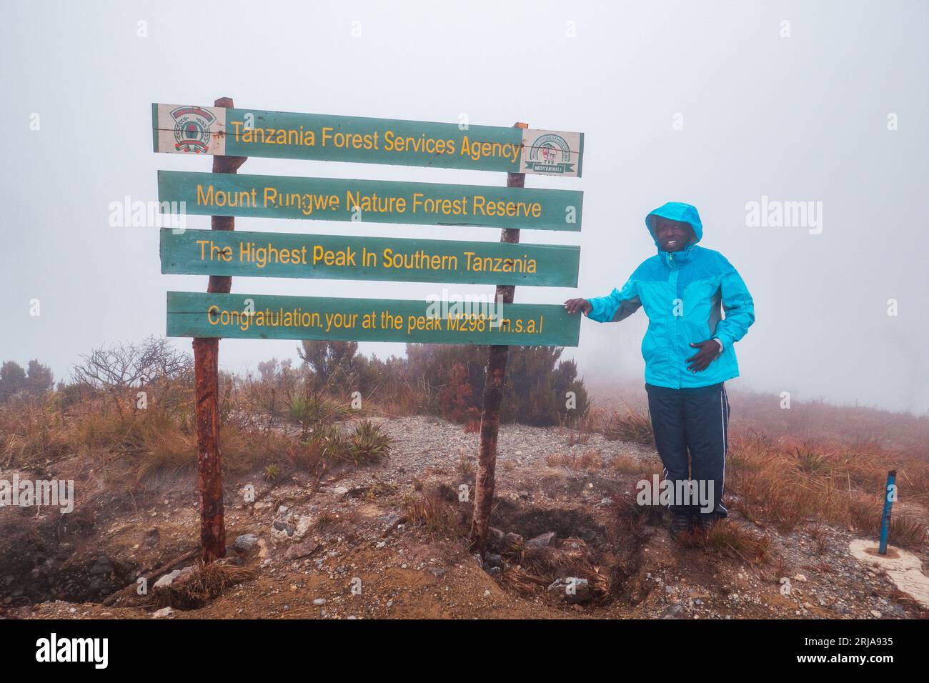 A hiker wearing rain clothes standing next to a sign post at the summit ...