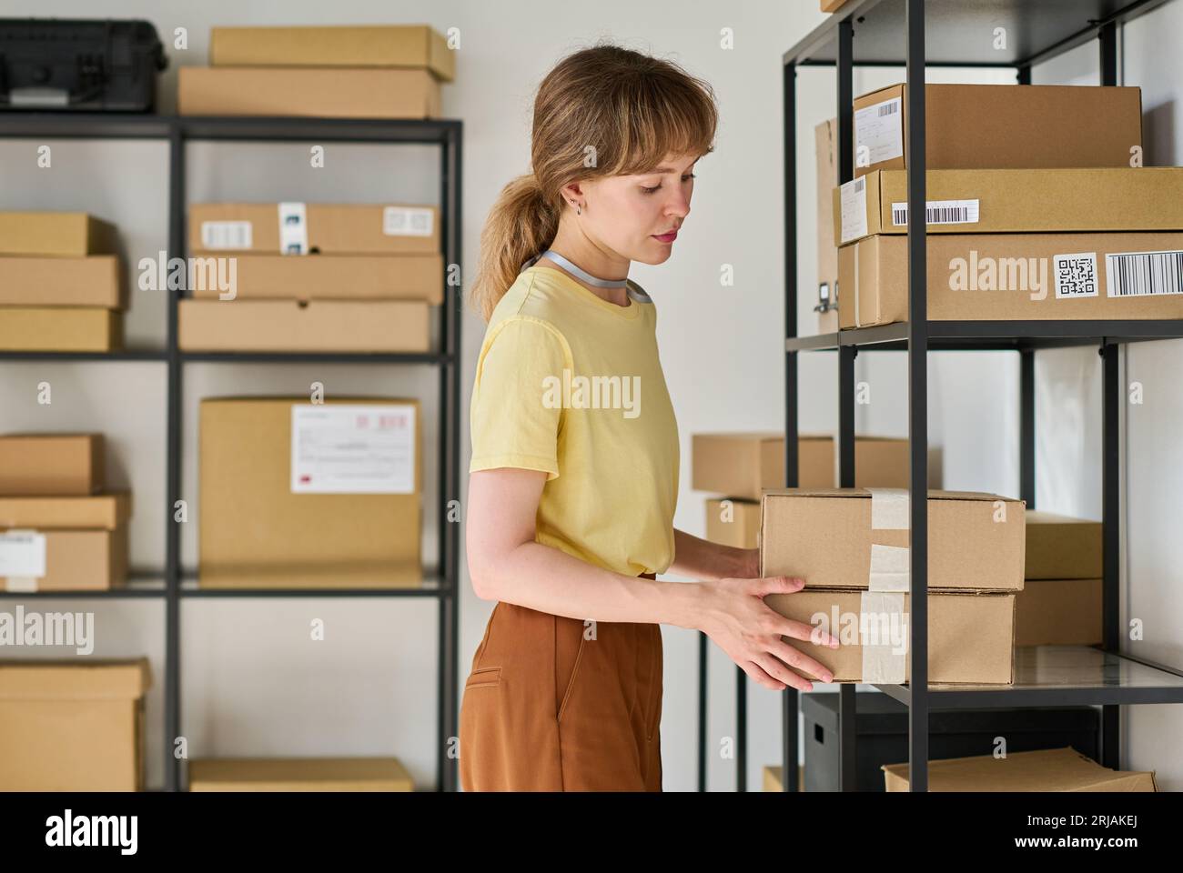 Side view of young woman in casualwear putting stack of boxes on shelf ...