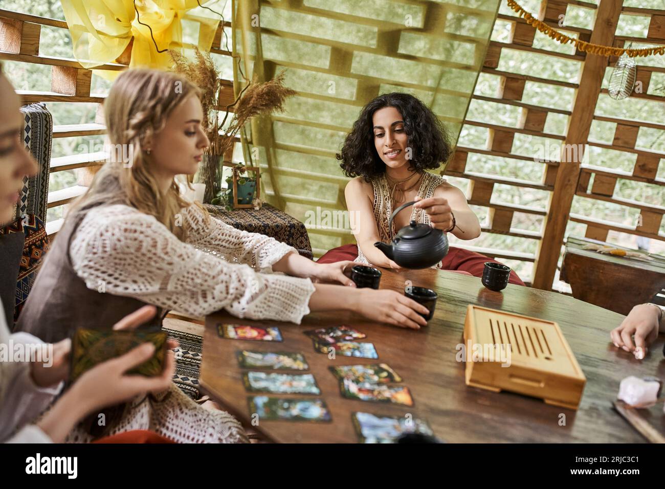 stylish multiracial woman pouring tea while young girlfriends reading ...