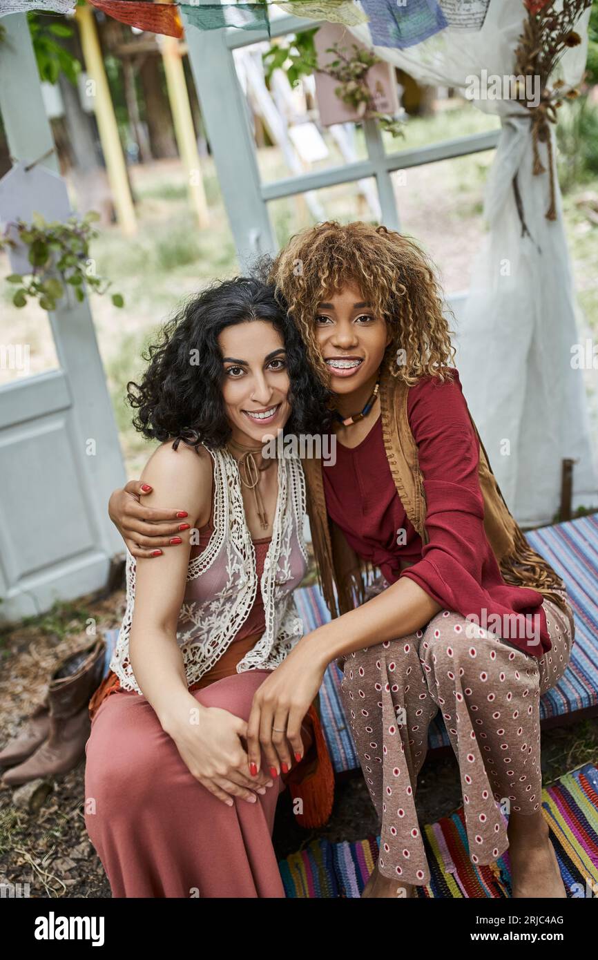smiling interracial girlfriends in boho outfits hugging and looking at ...