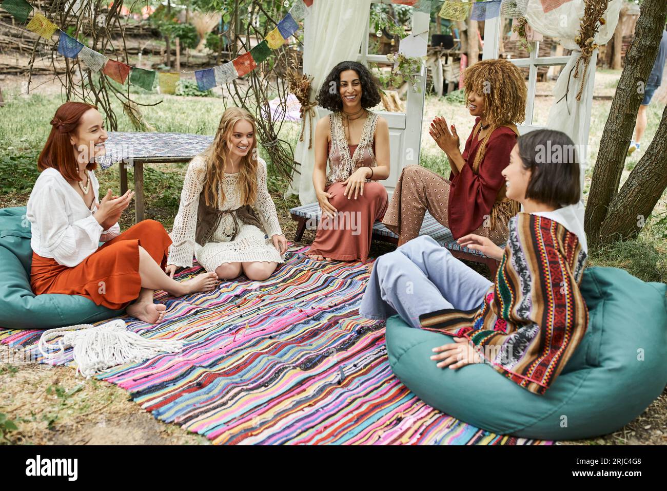 laughing girlfriends in boho style outfit talking on colorful blanket ...