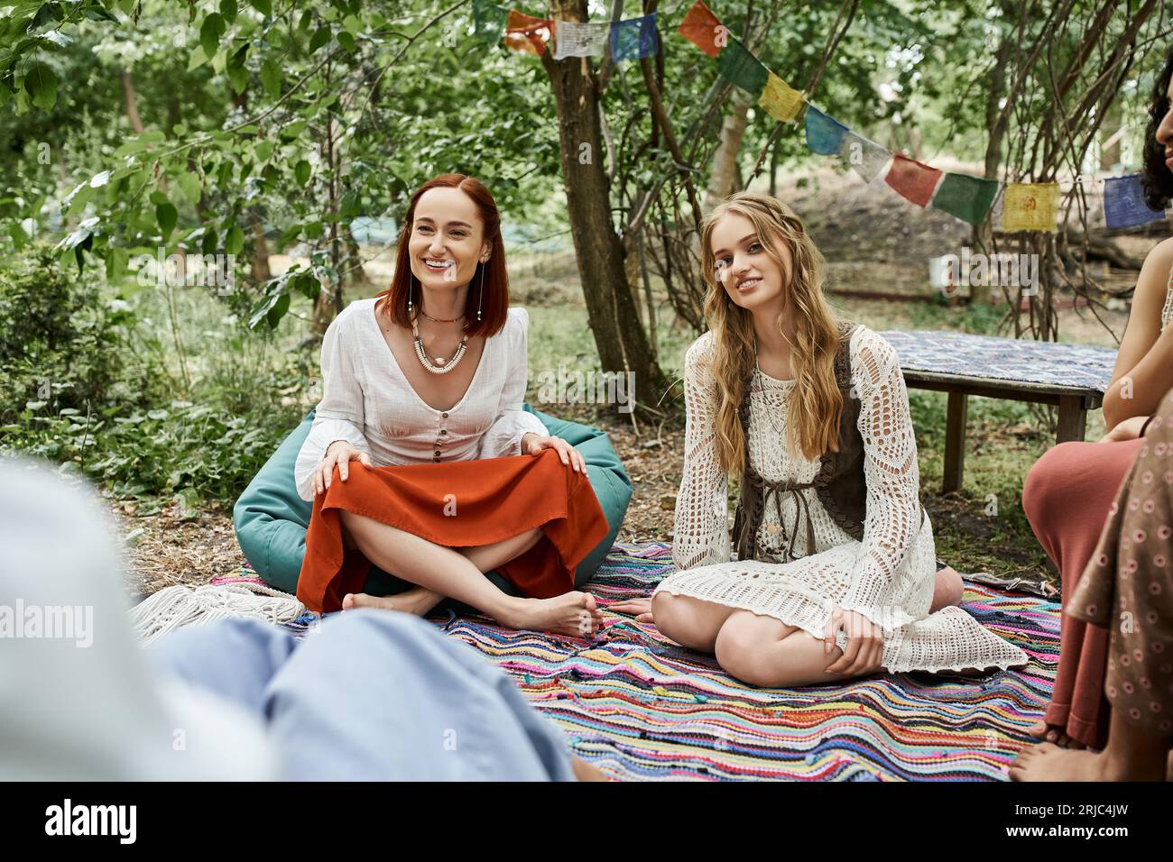 smiling and trendy women in boho styled outfits sitting on carpet ...