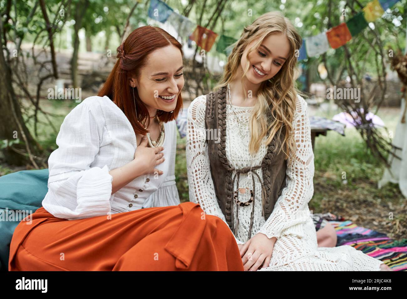 laughing woman in boho styled clothes sitting near friend on carpet on ...