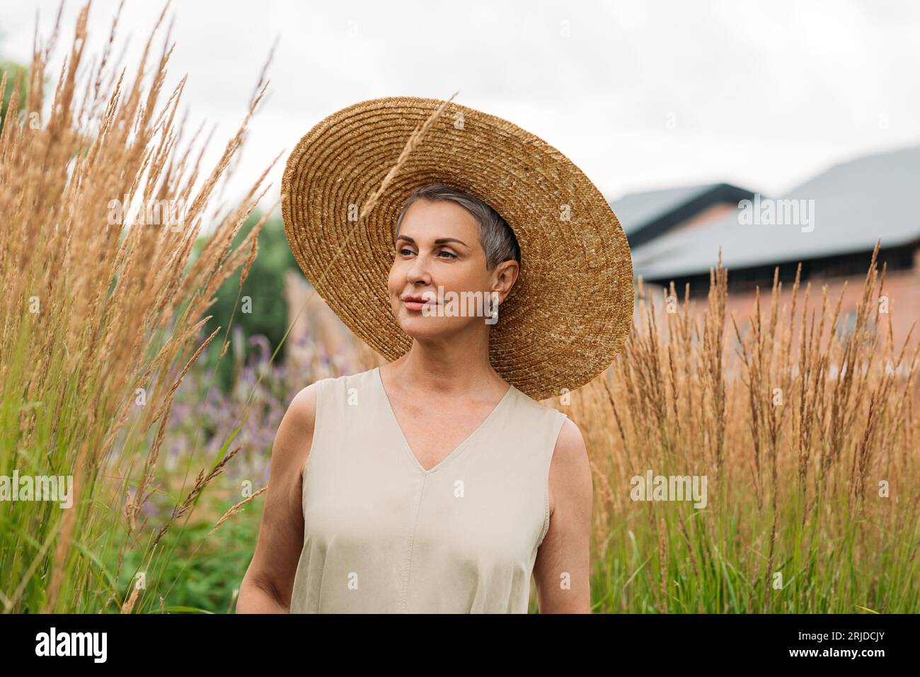 Aged woman with grey hair wearing a big straw hat walking on the wheat ...