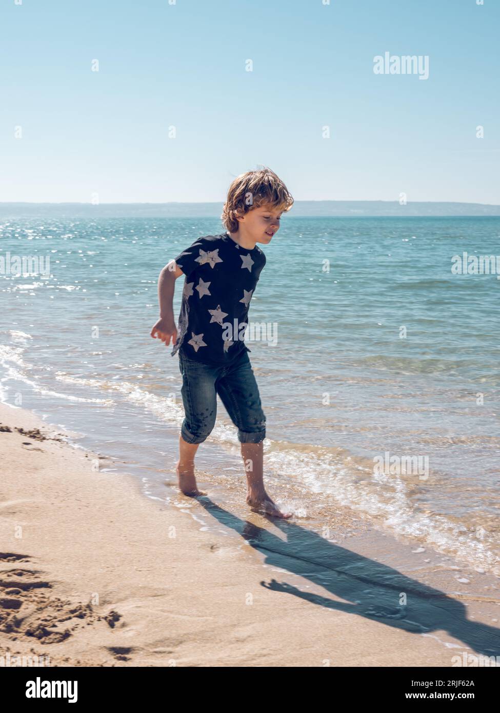 Blond delightful child walking in shallow water of waving sea in sunshine under cloudless blue ...