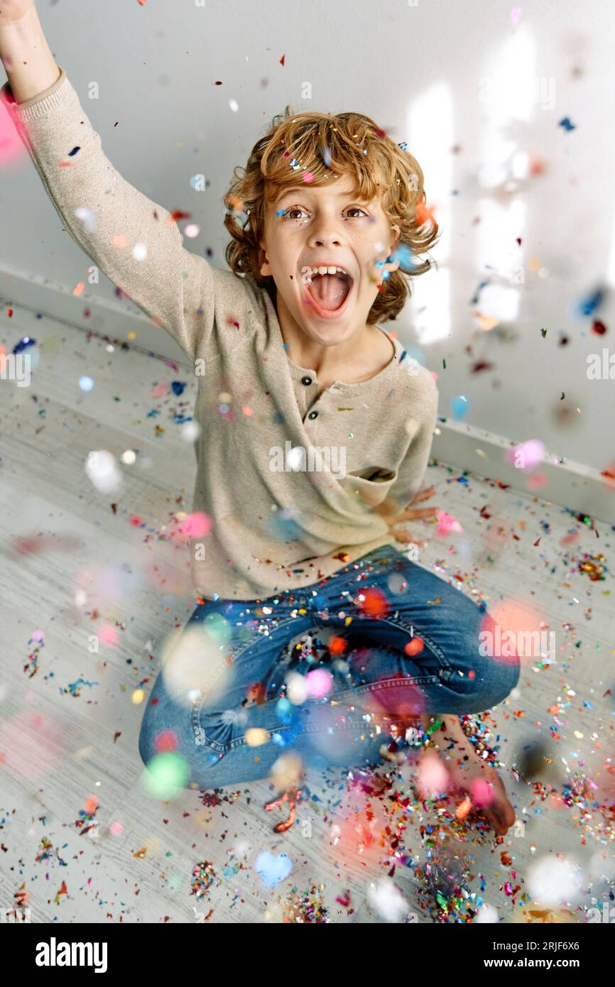 High angle of excited curly haired boy sitting on floor and throwing ...