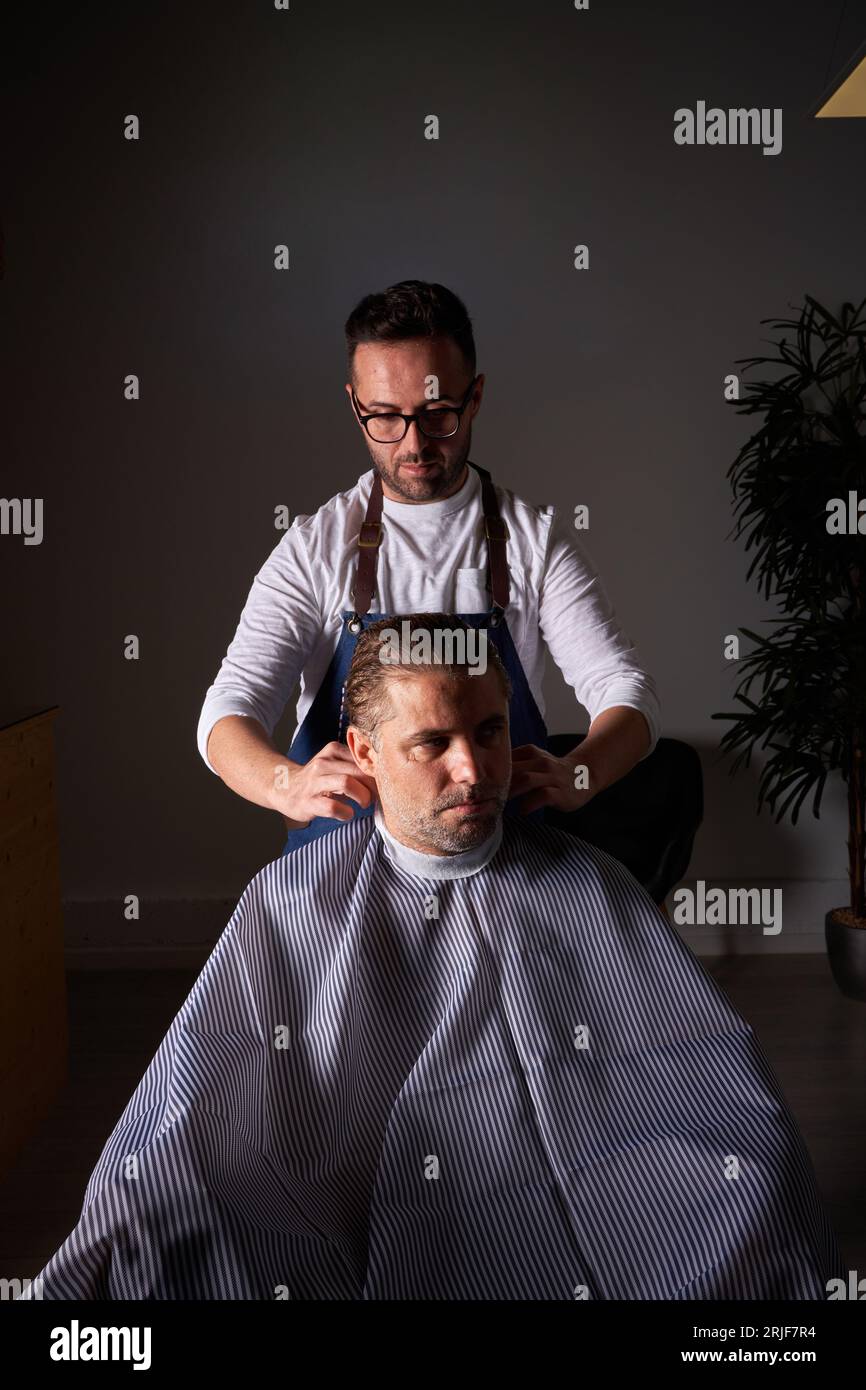 Professional male barber in apron and eyeglasses cutting hair of ...