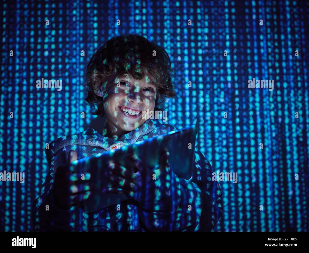 Smiling kid hacking digital tablet while standing in dark room with ...
