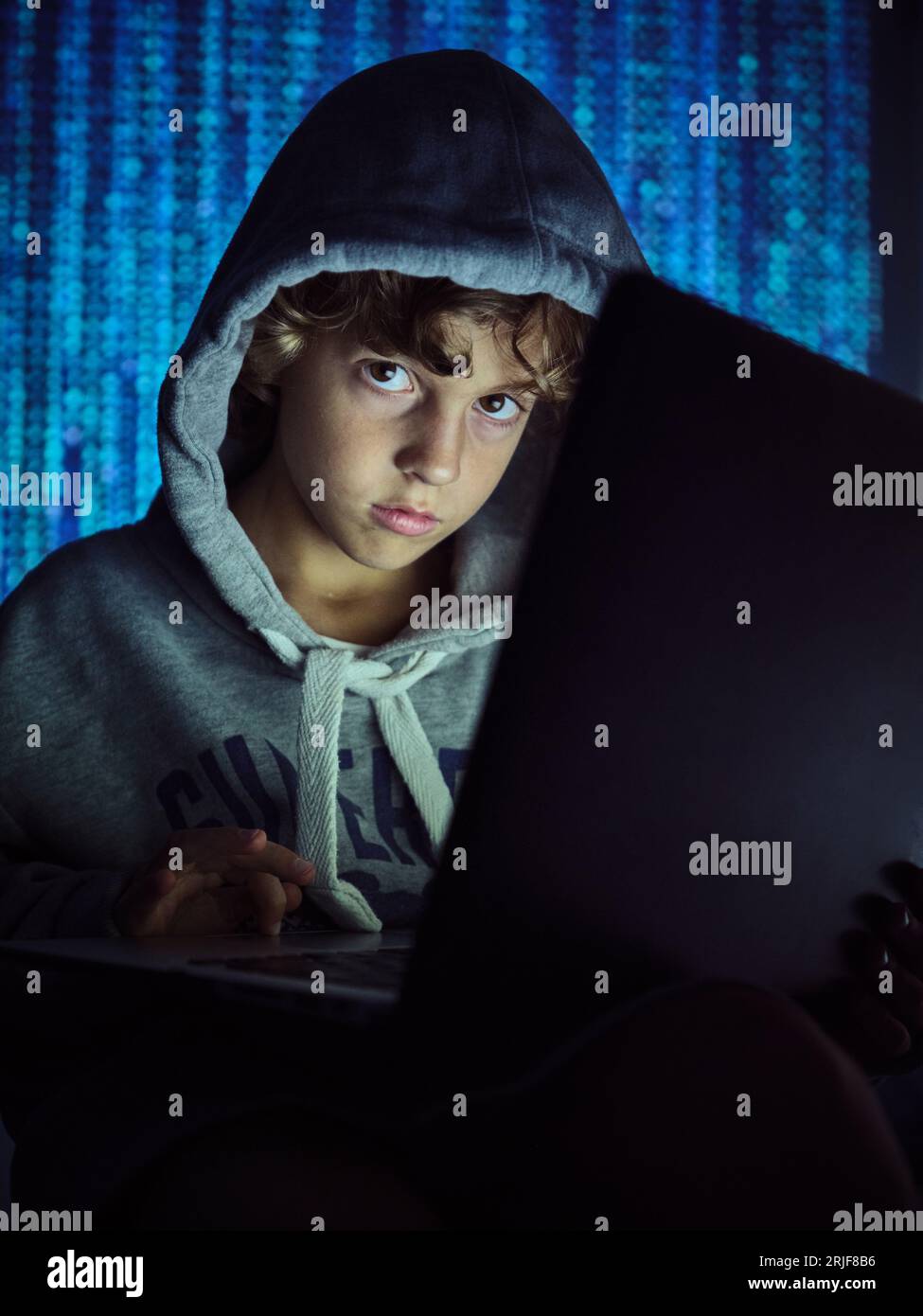 Serious hacker kid in hoodie typing on laptop on blurred binary code ...