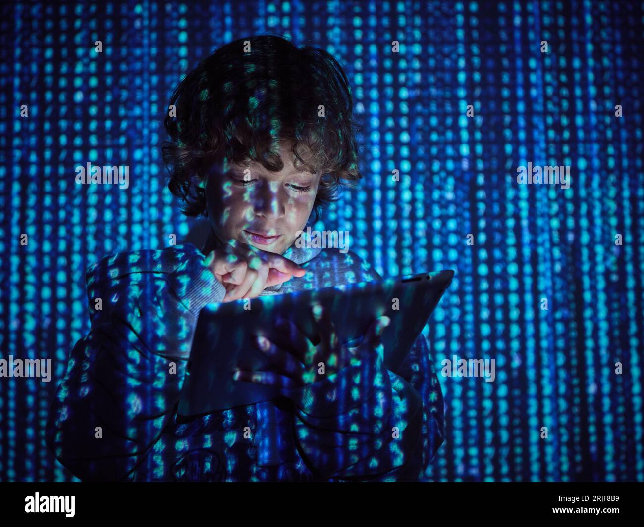 Teen boy pressing button on tablet to start hack while standing in dark room with code symbols ...