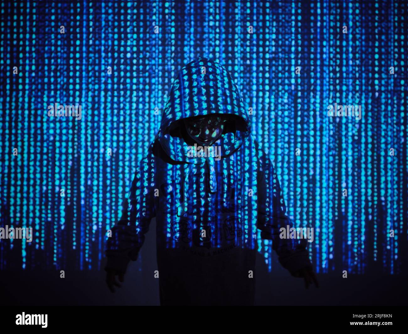 Anonymous hacker boy in hoodie standing in projection of blue binary code in dark room Stock ...