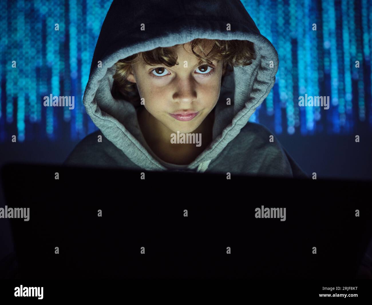 Crop kid sitting at laptop and hacking while looking at camera on blurred binary code background ...