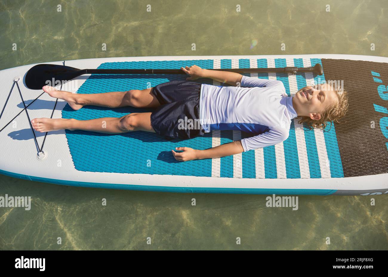From above full body of happy boy in activewear lying on paddleboard ...