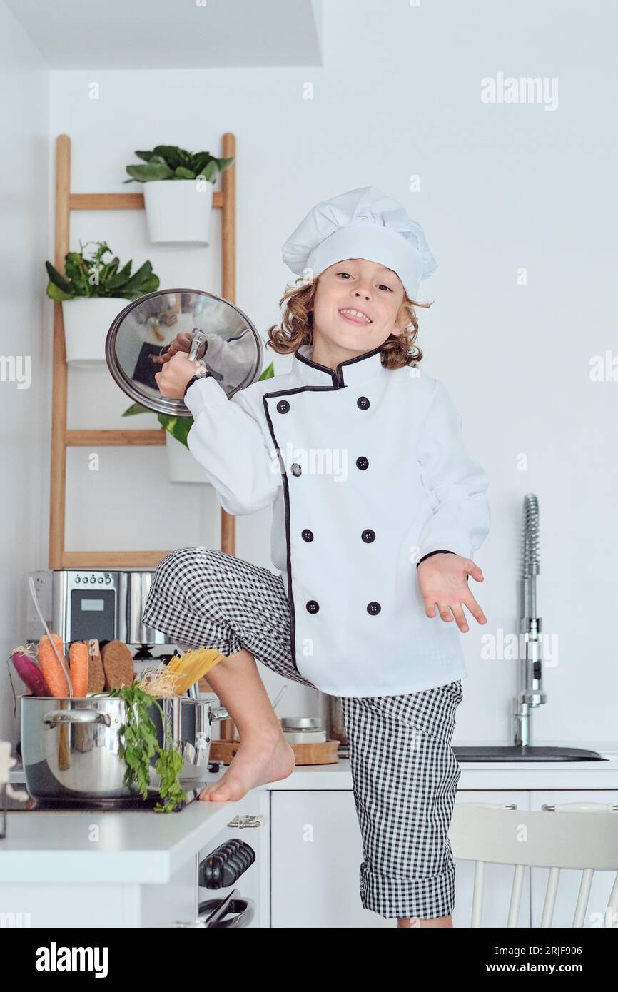 Positive smiling kid in chef hat and tunic standing near stove with pan ...