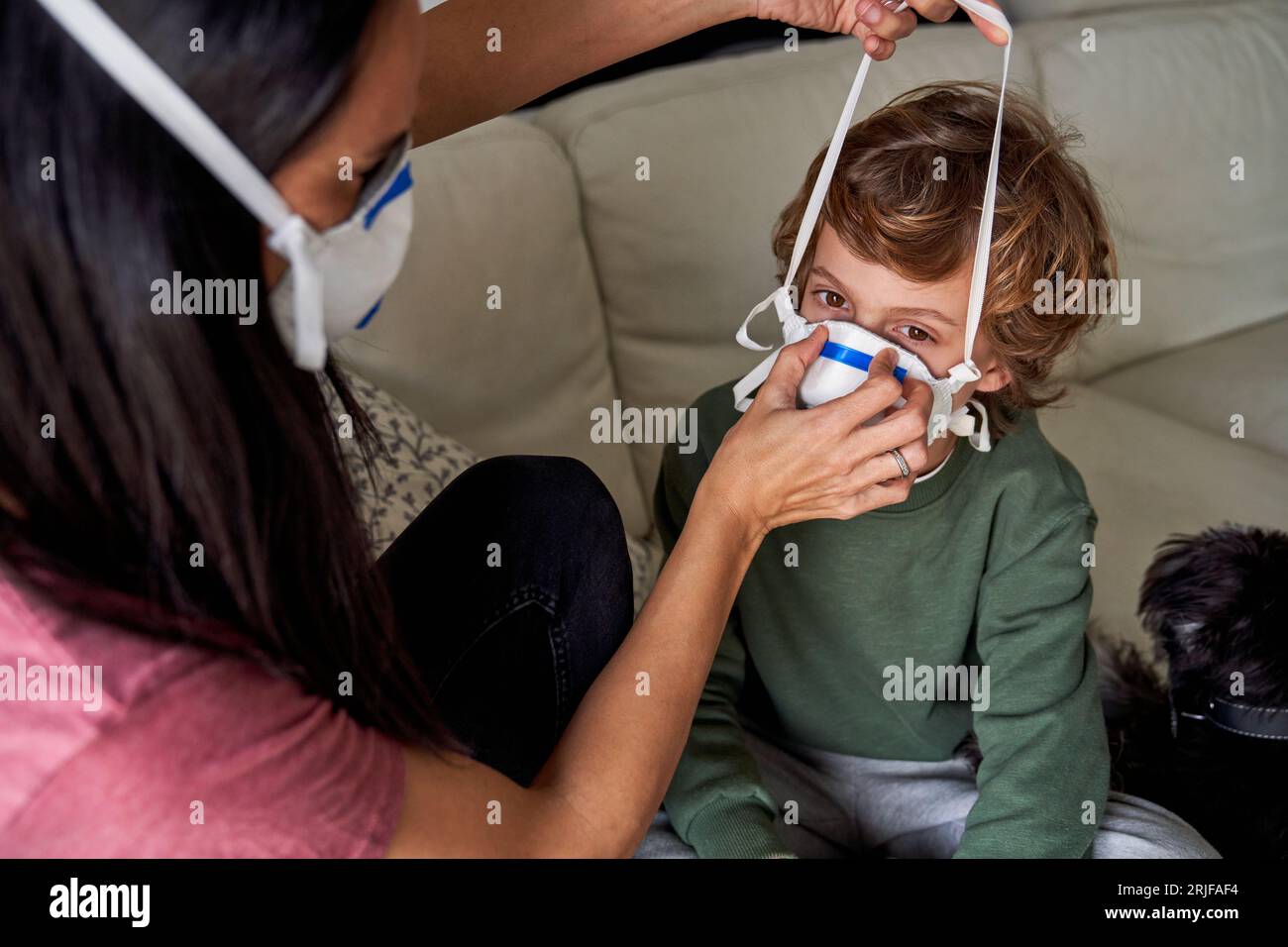 High angle of blond boy having protective mask put on face by crop mom ...