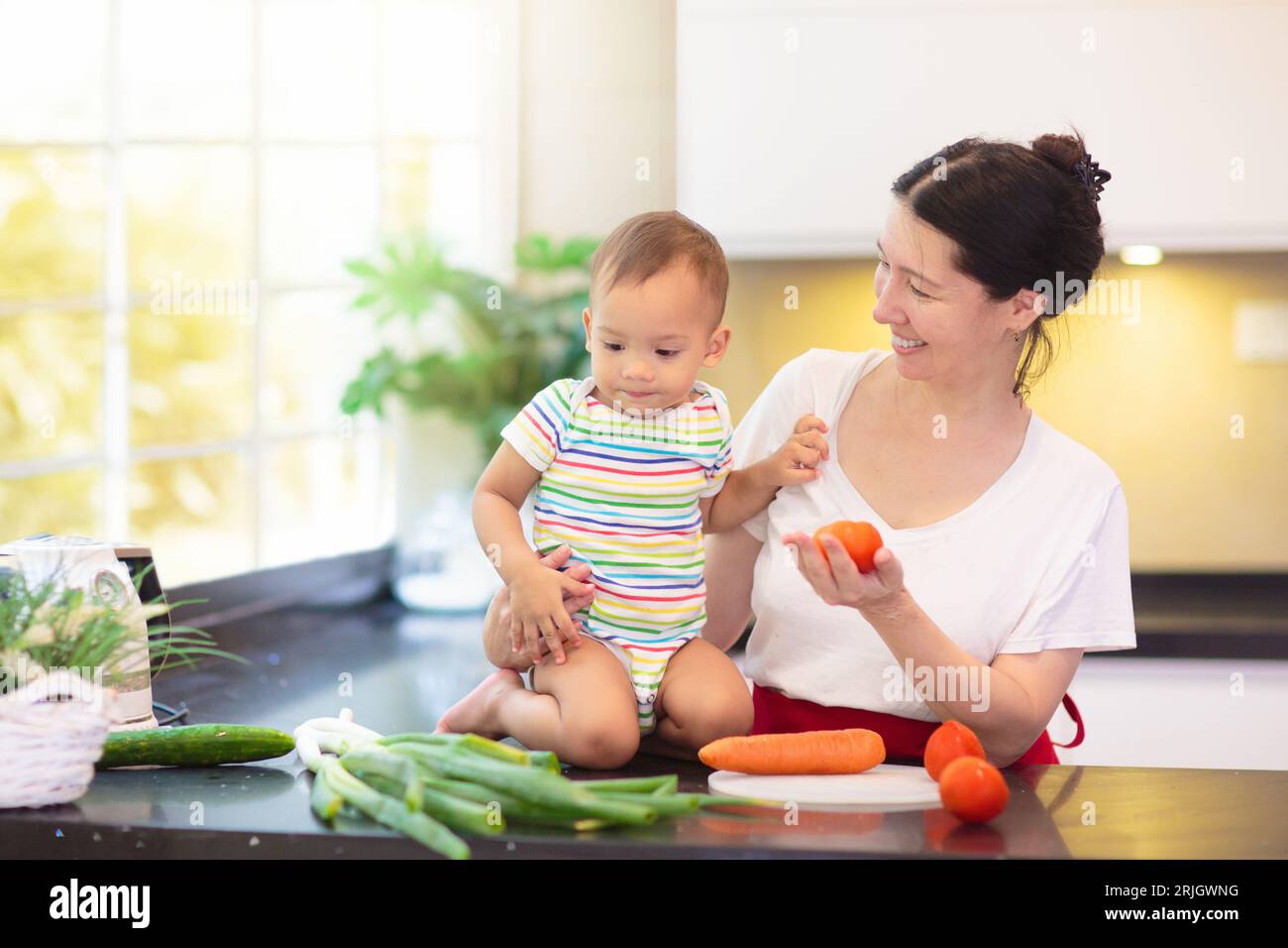 Mother and baby boy in white sunny kitchen. Young Asian mom cooking ...