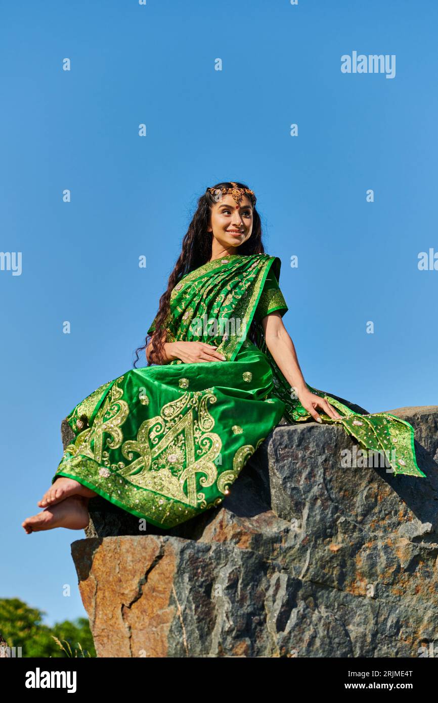 carefree barefoot indian woman in stylish green sari sitting on stone ...