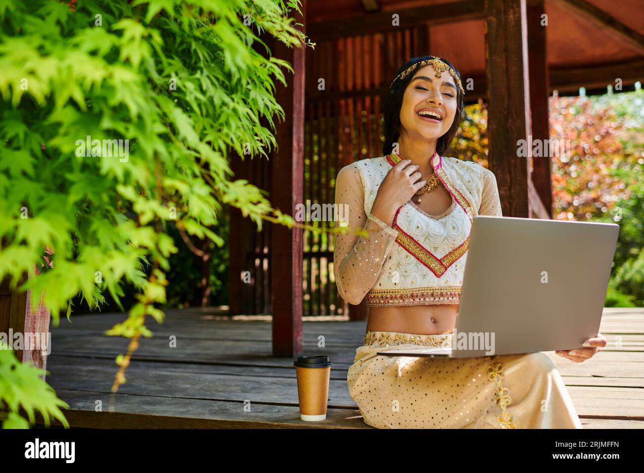 laughing indian woman sitting with laptop near coffee to go in wooden ...