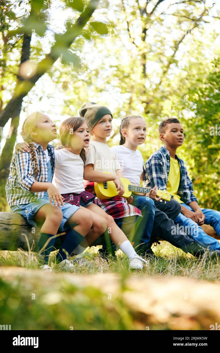 Happy children sitting on log in forest on sunny day, playing guitar ...