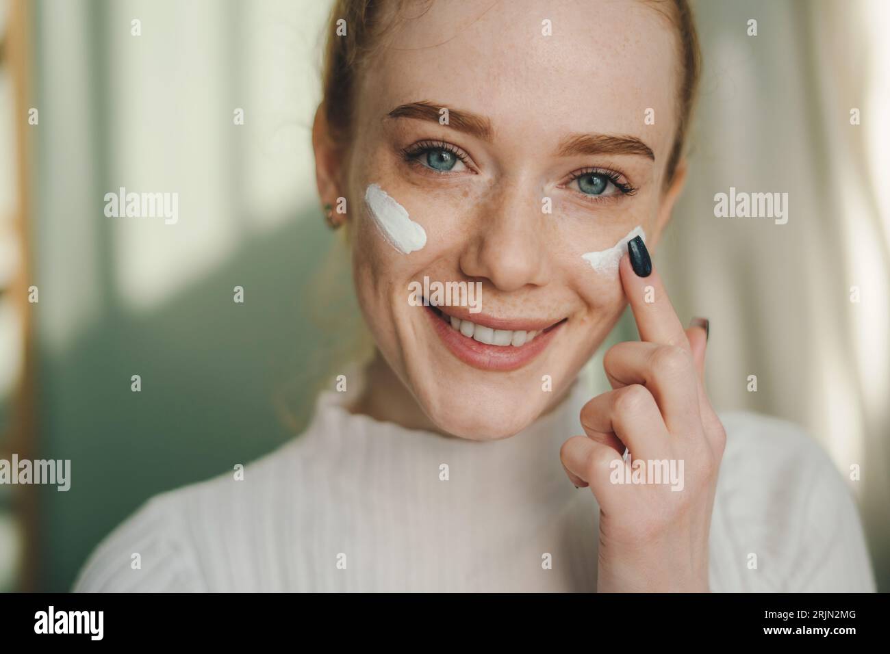 Redhead young woman putting moisturizing cream on face with freckles ...