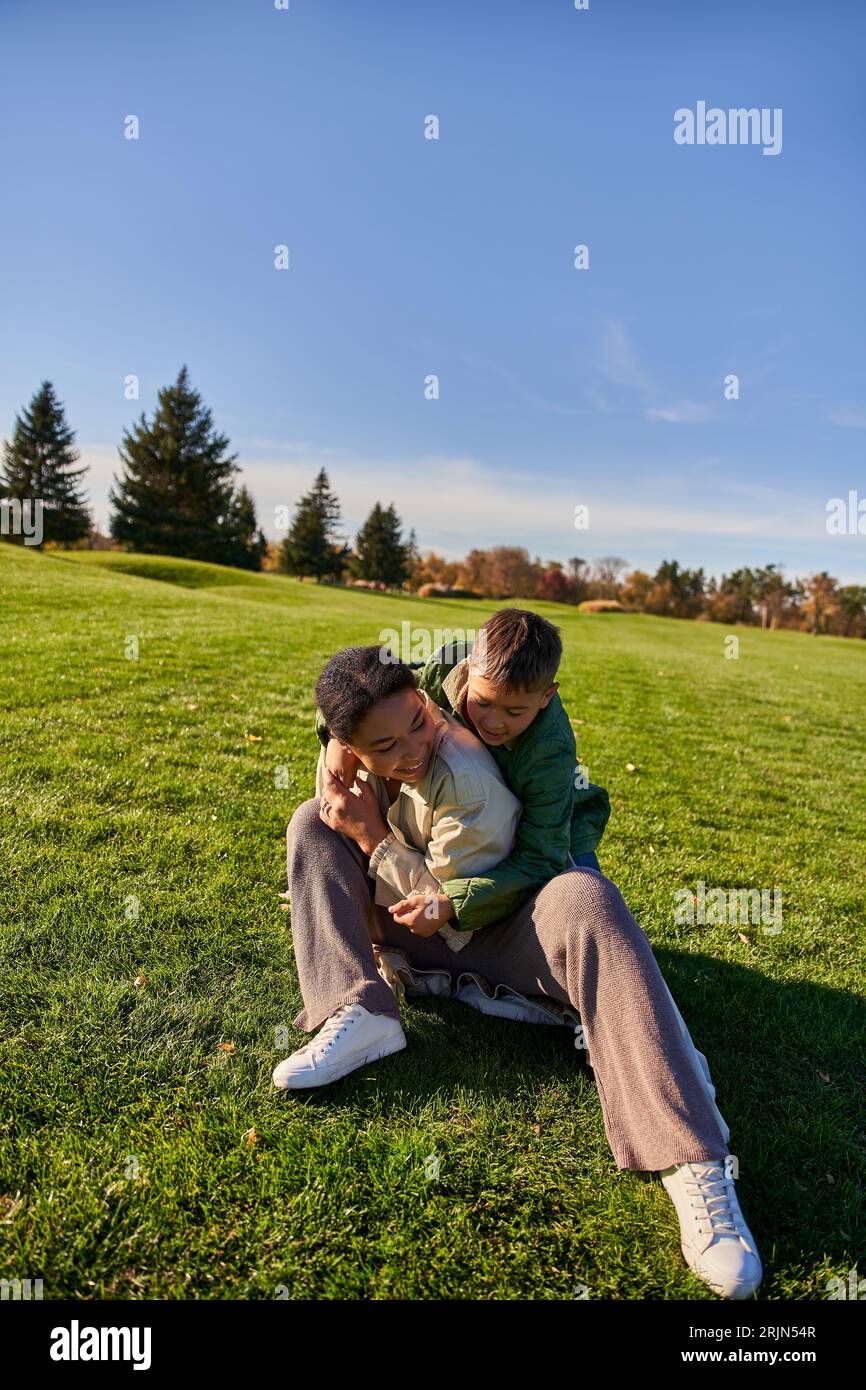 joyful mother and son sitting on grass, sunny day, autumn, playful ...