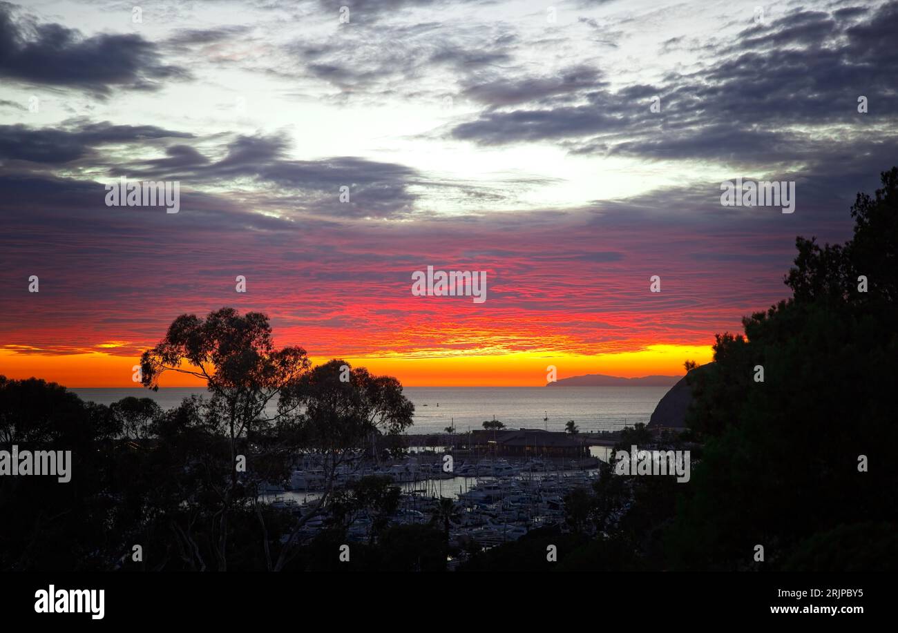A picturesque view of the sun setting over a harbor, silhouetting the masts and hulls in the ...